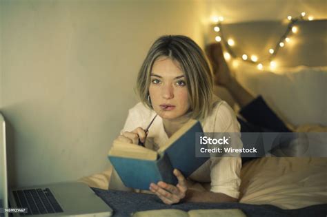 안경을 쓴 사려 깊은 여성의 얕은 초점은 노트북이 있는 침대에서 책을 읽고 있습니다 갈란드 장식품에 대한 스톡 사진 및 기타