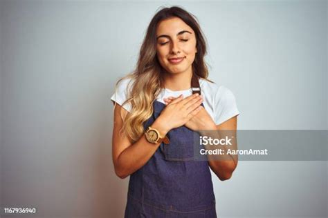 회색 고립 된 배경위에 앞치마를 입은 젊은 아름다운 여성은 닫힌 눈과 얼굴에 감사하는 제스처로 가슴에 손을 대고 미소 짓고 있습니다 건강 개념 감사에 대한 스톡 사진 및