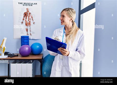 Young Blonde Woman Wearing Physiotherpist Uniform Writing On Checklist At Physiotherapy Clinic