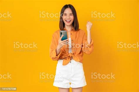 헤드폰을 들고 주먹으로 스마트폰을 들고 노란 배경에서 성공을 거둔 승리를 축하하는 쾌활한 젊은 아시아 여성 스튜디오 촬영에 대한 스톡 사진 및 기타 이미지 Istock