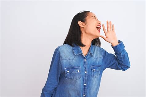 고립된 흰색 배경 위에 서있는 데님 셔츠를 입은 젊은 아름다운 중국 여성이 입에 손을 대고 큰 소리로 외치고 비명을 지르고 있습니다 통신 개념입니다 갈색 머리에 대한 스톡