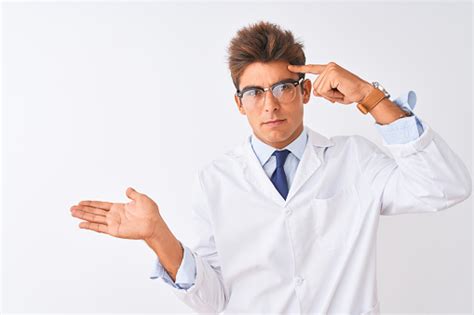고립 된 흰색 배경 위에 안경과 코트를 입은 젊은 잘생긴 Sciencist 남자는 혼란스럽고 열린 손바닥으로 카피 공간을 보여주고 이마를 가리키는 손가락을 괴롭혔습니다