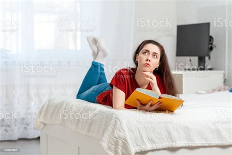 한 젊은 백인 여성이 침대에 누워 서서 책을 사려 깊게 읽고 있습니다 공간을 복사합니다 배경에 방의 흰색 인테리어 교육과 세계 도서의 날의 개념 갈색 머리에 대한 스톡 사진