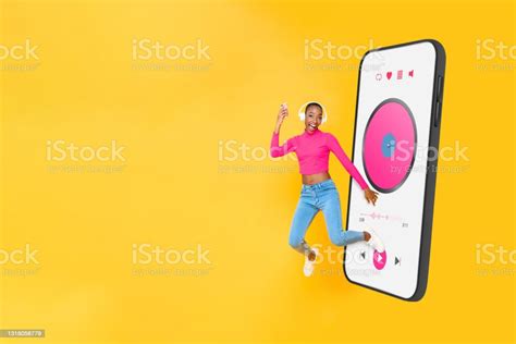 화려한 노란색 배경에 고립 된 스마트 폰에서 응용 프로그램에서 음악에 헤드폰 목록을 착용 점프 행복 한 활기찬 아프리카 계 미국인 여성의 전체 길이 초상화 십대에 대한 스톡