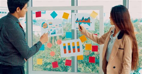 비즈니스 사람들은 사무실의 프로젝트 계획 보드에서 일하고 동료 친구와 대화하여 프로젝트 개발을 분석합니다 그들은 유리 벽에 게시 된 스티커 메모를 사용하여 정리합니다