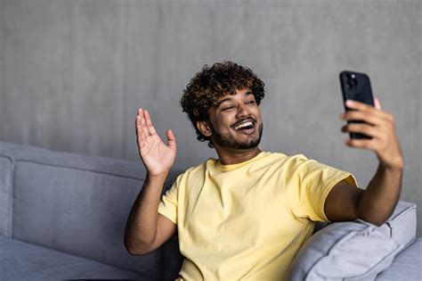 만나서 반갑습니다 가상 통신을 위해 스마트폰 앱을 사용하여 캐주얼한 옷을 입은 쾌활한 인도 남자 손에 전화기를 들고 웹캠을 향해 손을 흔들고 소파에 누워 화상 통화를 들고