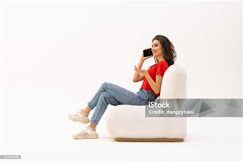 웃고 있는 젊은 여자가 전화로 대화를 나누고 하얀 스튜디오 벽에 아늑한 의자에 앉아 있다 스마트폰으로 고객이나 친구와 이야기하는 아름다운 백인 여성 말하기에 대한 스톡 사진