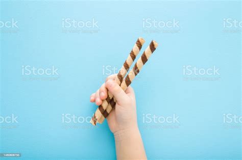 밝은 파란색 테이블 배경에 두 개의 줄무늬 초콜릿 웨이퍼 스틱을 들고 있는 어린 아이 파스텔 색상 근접 촬영 어린이 달콤한 간식 하향식 보기 끈적거리는에 대한 스톡 사진 및