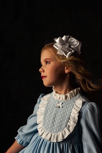 검은 배경에 사랑스러운 작은 인형 덮개 소녀가 시선을 돌리고 있다 파란 드레스를 입고 빛과 그림자로 격리된 머리에 하얀 활을 얹은 이미지의 사랑스러운 아이 스튜디오 패션