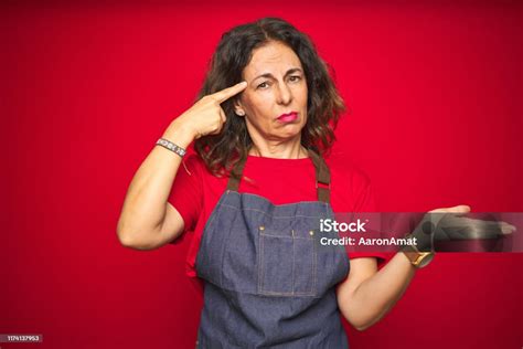 빨간 고립 된 배경 위에 앞치마 유니폼을 입고 중년 노인 여성은 카피 공간을 보여주는 열린 손바닥과 이마에 손가락을 가리키는 짜증 그것에 대해서 생각해 봐 앞치마에 대한