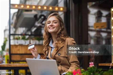 카페 테라스에서 야외에서 노트북과 함께 앉아 있는 신용 카드를 손에 들고 카메라를 들고 행복하게 트렌치코트를 입은 젊은 즐거운 여성 16 17 살에 대한 스톡 사진 및 기타