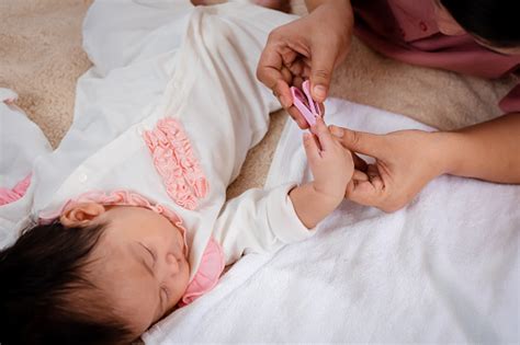 아기 소녀는 갈색 침대에서 편안하게 자고 어머니는 아기가 잠들 시간을 갖고 잠자는 아기의 손톱을 조심스럽게 자르기 위해 클리퍼를 가져 와서 아기가 느끼지 않도록했습니다 2