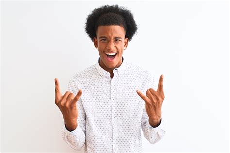 고립 된 흰색 배경 위에 서있는 우아한 셔츠를 입은 젊은 아프리카 계 미국인 남성이 미친 표정으로 손을 들고 바위 기호를하고 있습니다 음악 스타 무거운 개념 개념에 대한