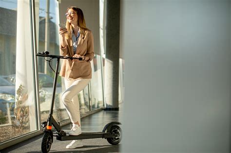 전기 스쿠터와 사무실 창 옆에 서서 휴대 전화를 사용하여 젊은 여자 20 24세에 대한 스톡 사진 및 기타 이미지 20 24