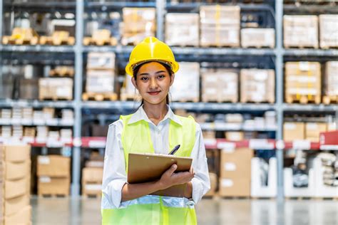 Asian Woman Warehouse Worker In Uniform Doing Stocktaking Of Products Management In Cardboard