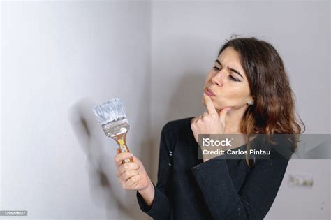 의심스럽고 사려 깊은 얼굴로 하얀 벽을 바라보는 페인트 브러시를 들고 있는 젊은 여성 30 34세에 대한 스톡 사진 및 기타 이미지 30 34세 Diy 가사 집안일