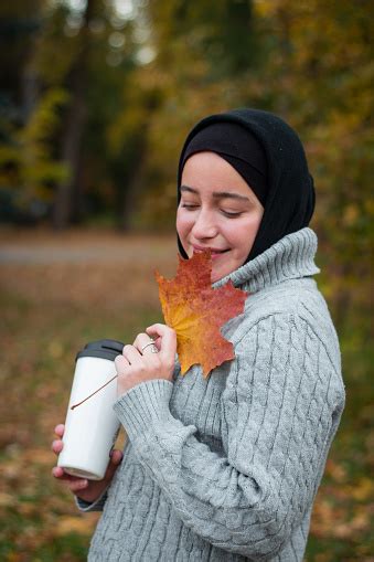 커피 한 잔과 노란 가을 잎을 가진 회색 스웨터를 입은 젊은 무슬림 여성 가을 공원 여성스러움과 미소 20 24세에 대한 스톡