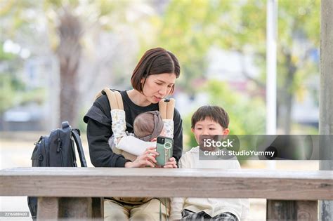 봄 후쿠오카의 한 공원에서 30대 엄마가 3살짜리 아들과 6개월 된 딸아이를 데리고 벤치에 앉아 있다 그들은 함께 스마트폰을 보며 차분하고 연결된 가족 시간을 공유합니다 2