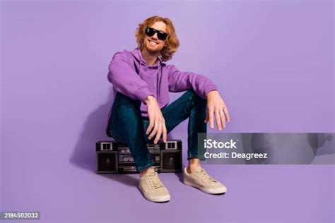시원한 수염 빨간 머리 남자 앉아 듣다 붐 박스 착용 선글라스 까마귀 청바지 신발 보라색 배경에 고립 된 전신 사진 30 34