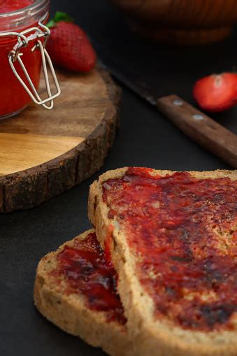 나무 도마에 유리 병입 항아리에 집에서 만든 딸기 잼의 클로즈업 이미지 녹색 잎 잼 작은 술 칼 검은 배경 전경에 초점을 맞춘 흰색 토스트 신선한 빨간 딸기의 전체 신선한