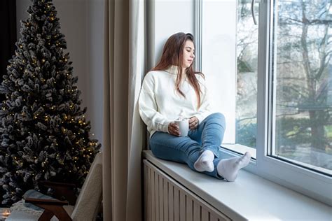 따뜻한 흰색 겨울 스웨터를 입은 여자는 크리스마스 이브에 집에서 창턱에 앉아 마시멜로가 든 컵을 들고 있다 12월에 대한 스톡 사진 및 기타 이미지 Istock