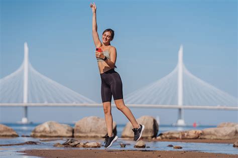 운동복을 입은 젊은 아름다운 웃는 갈색 머리 여자가 해변에서 점프하며 다리와 만을 배경으로 화상 통화를 하고 있다 예쁜 이탈리아 적합 여성은 공원에서 아침 야외 운동 점프를