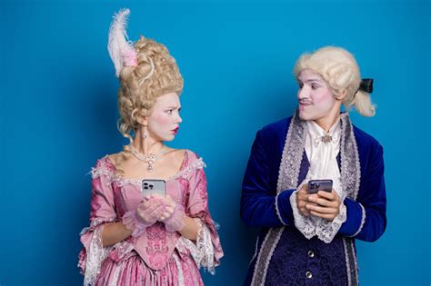 바로크 양식의 의상을 입은 기발한 커플이 파란색 배경에서 스마트폰으로 문자를 보내며 과거의 우아함과 현대 기술이 조화를 이루고