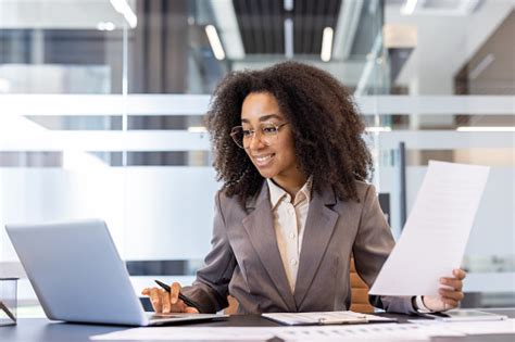 미소 짓는 젊은 아프리카계 미국인 여성이 사무실 책상에 앉아 문서를 들고 노트북 화면을 보고 있습니다 검사 보기에 대한 스톡 사진 및 기타 이미지 Istock