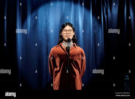 Young Chinese Female Comedian In Brown Shirt Standing On Stage With Blue Curtains And Speaking