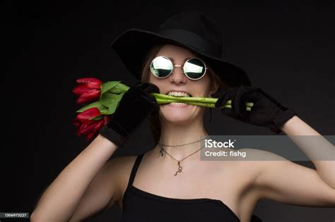 모자와 선글라스를 입은 젊은 여성의 스튜디오 초상화가 그녀의 입에 빨간 튤립 꽃다발을 들고 검은 색 배경에 검은색에 대한 스톡