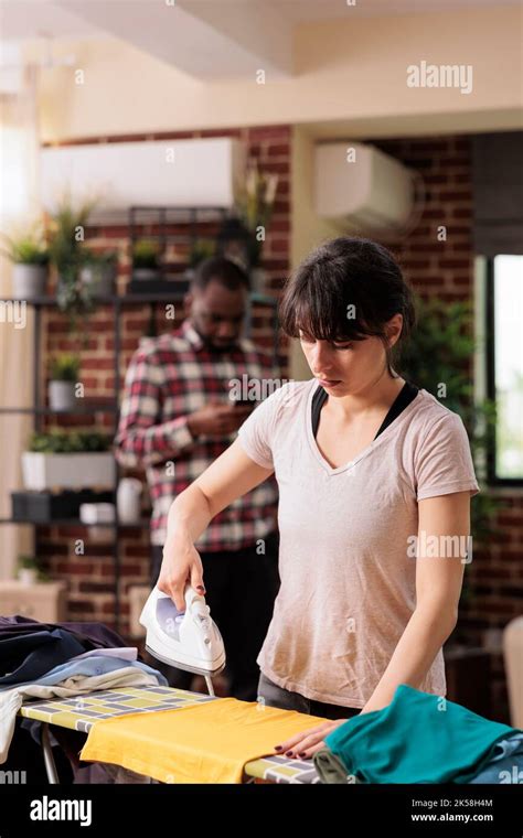 Woman Ironing Clothes At Home While Husband Distracted Using Mobile