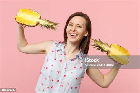 스튜디오에서 분홍색 파스텔 벽 배경에 고립 된 신선한 잘 익은 파인애플 과일의 Halfs를 들고 여름 옷에 젊은 여자를 웃는 사람들은 생생한 라이프 스타일 휴식 휴가