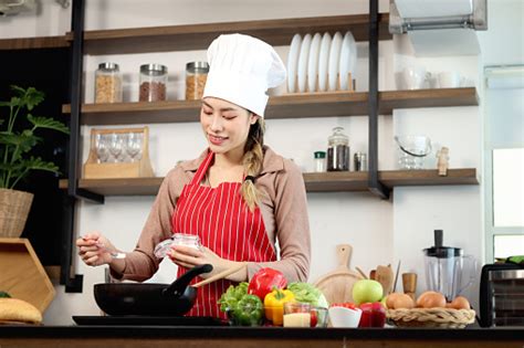 앞치마와 요리사 모자를 쓰고 행복한 미소를 짓고 있는 아름다운 아시아 여성 주방에서 맛있는 튀긴 음식을 요리하기 위해 팬에 조미료를 추가하고 여성 요리사가 맛있는 건강