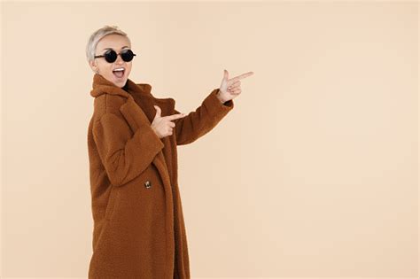 금발의 짧은 머리를 가진 힙스터 여성은 베이지 색 배경 위에 고립 된 코트와 선글라스 포인트 손가락을 착용합니다 베이지에 대한 스톡 사진 및 기타 이미지 Istock