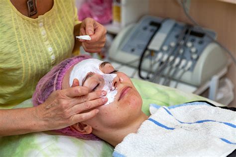 숙련 된 미용 학자는 얼굴 클렌징 및 미용 치료 중에 소파에 놓여 있는 어린 소녀의 얼굴에 흰 크림 마스크를 적용 합니다 내각 미용사 피부 건강과 청소년 거실에 대한 스톡