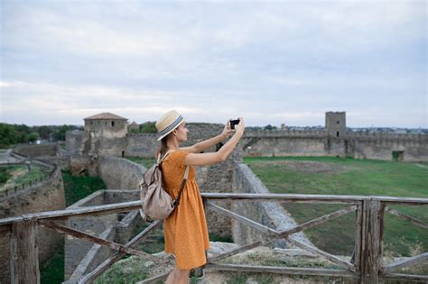 밀짚 모자와 드레스를 입은 젊은 여성이 고대 성이나 요새 벽에 서서 맑고 푸른 하늘을 배경으로 셀카를 찍습니다 역사적인 건축물과 조화를 이루는 경치 좋은 전망 30 34세에