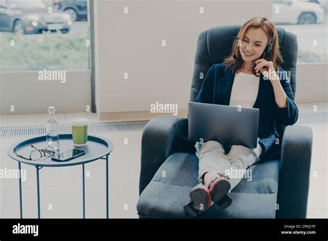 Successful Redhead Woman Entrepreneur Talking Online With Coworkers Sitting On Armchair In