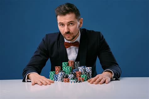 검은 양복을 입은 남자가 파란색 스튜디오 배경에서 포즈를 취하고 있는 컬러 칩더미가 있는 흰색 테이블에 앉아 있다 도박 포커 카지노 클로즈업 갈색 머리에 대한 스톡 사진 및