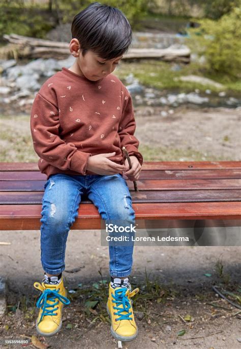 갈색 청바지 작업복과 노란 부츠를 입고 시냇물 옆의 벤치에 앉아 손에 나뭇 가지를 들고 내려다 보는 라티노 소년을 당황스럽게 만들었습니다 가을 날씨 세로 가을에 대한 스톡