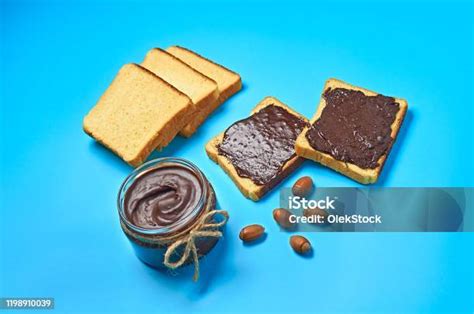 녹은 초콜릿이나 크림으로 가득 찬 유리 항아리는 바닐라 빵과 헤이즐넛 조각 근처에 부엌의 파란색 책상에 놓여 있습니다 영양가 있는 아침 식사 또는 점심 식사의 개념 가득