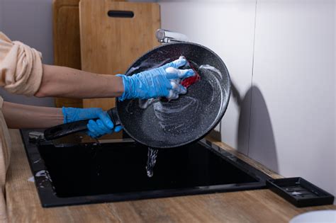 천연 목재 요소를 배경으로 하고 위생을 위한 보호 장갑을 낀 현대적인 주방 환경에서 비누와 물로 검은색 붙지 않는 팬을 청소하는 사람 가정 주방에 대한 스톡 사진 및 기타