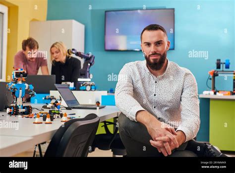 A Man Sitting In A Robotics Laboratory While His Colleagues In The