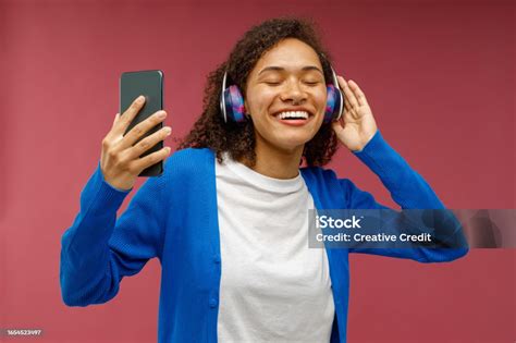 이어폰을 끼고 춤을 추고 빨간 스튜디오 배경에서 음악을 듣는 쾌활한 여자 25 29세에 대한 스톡 사진 및 기타 이미지 25 29세 Brand Name Smart