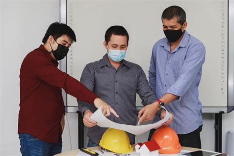 사무실에서 함께 일하는 보호 마스크를 착용한 건축가와 엔지니어의 자신감 있는 팀 그들은 책상에서 새로운 시작 프로젝트에 대해 논의합니다 건축 개념 사회적 거리 연습은 코로나