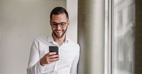 젊은 잘 생긴 웃는 우아한 남자는 실내에 서서 스마트 폰으로 메시지를 묶거나 읽습니다 사진 Unsplash의 메시지 이미지