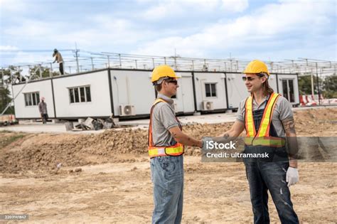 안전모와 안전조끼를 착용한 두 명의 건설 노동자가 건설 현장에서 악수를 하고 있는데 이는 모듈식 유닛을 배경으로 협업과 성공적인 팀워크를 상징합니다 2명에 대한 스톡 사진
