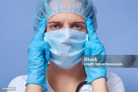 두통을 앓고있는 어린 소녀의 스튜디오 초상화 사원에 손을 잡고 의료 독감 마스크 고무 장갑과 일회용 모자를 착용하고 파란색 배경 위에 고립 된 포즈를 취합니다 힐르 케어
