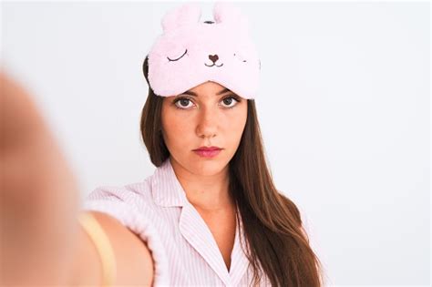 잠옷과 수면 마스크를 착용한 소녀는 스마트 한 얼굴에 자신감있는 표정으로 고립 된 흰색 배경 위에 카메라로 셀카를 만듭니다 갈색 머리에 대한 스톡 사진 및 기타 이미지