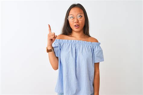 파란색 티셔츠와 안경을 쓰고 고립된 흰색 배경 위에 손가락을 가리키는 젊은 중국 여성이 성공적인 아이디어로 손가락을 가리킵니다 종료하고 행복 넘버 원 T 셔츠에 대한 스톡