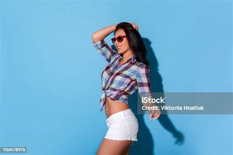 여름 시간입니다 반바지와 선글라스를 입은 여성은 파란 벽에 고립 된 맨 배를 들고 카메라를 만지는 머리를 만지는 장난스러운 측면